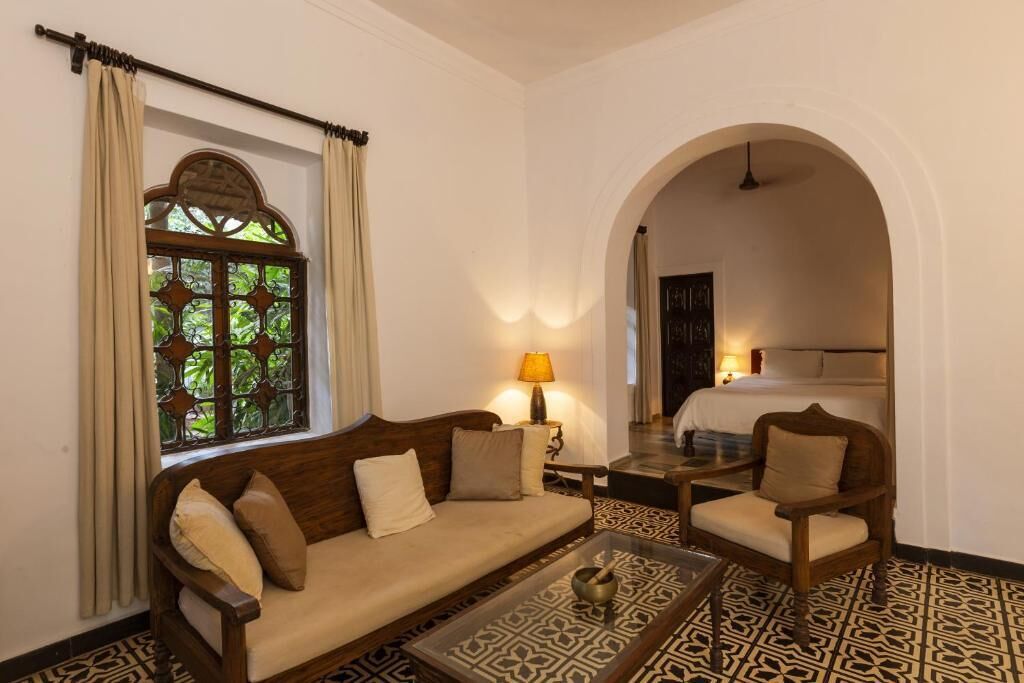 Quarto com sofá, poltrona e piso estampado. Há uma janela com cortinas e uma luminária em uma mesa lateral.