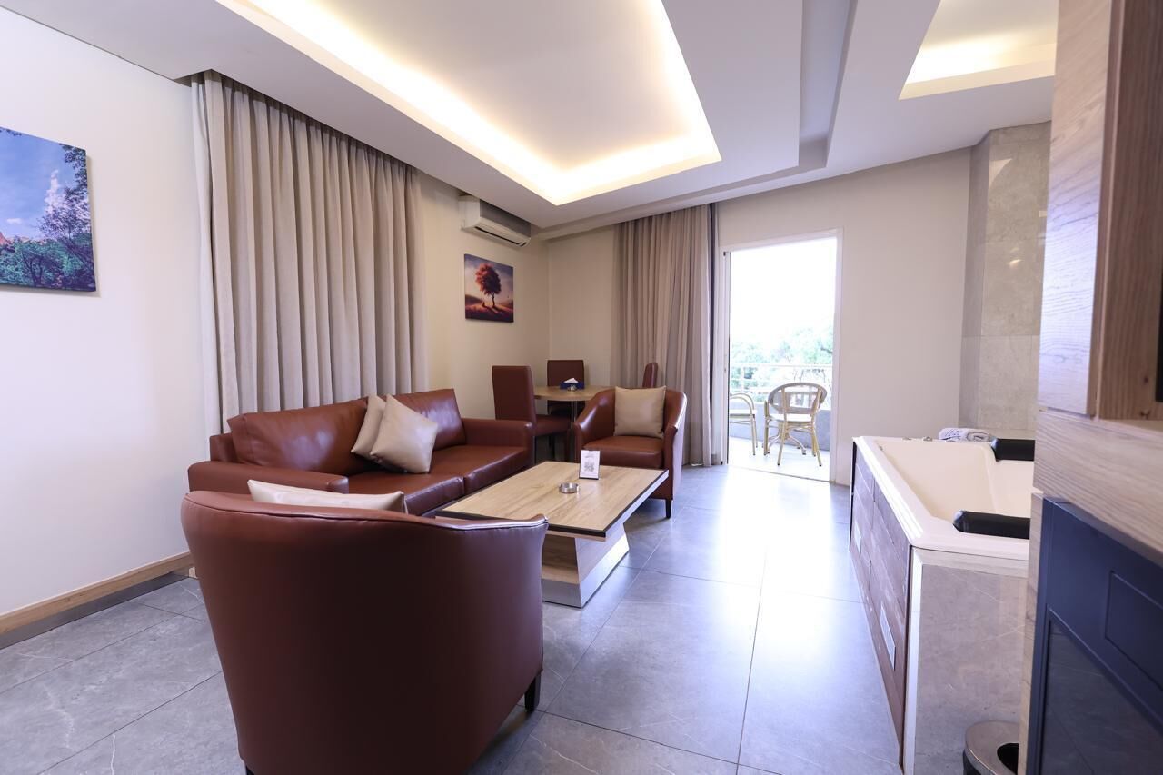 Quarto de hotel moderno com um sofá de couro marrom, uma mesa de centro de madeira e uma pequena área de refeições com uma mesa e cadeiras brancas.