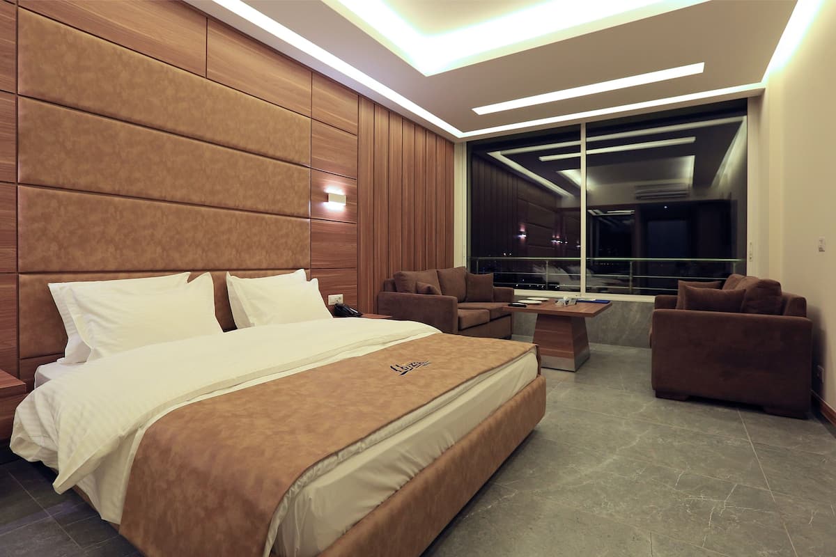 Quarto de hotel moderno com uma cama grande, duas poltronas, uma mesinha e vista para a cidade por uma janela ampla.