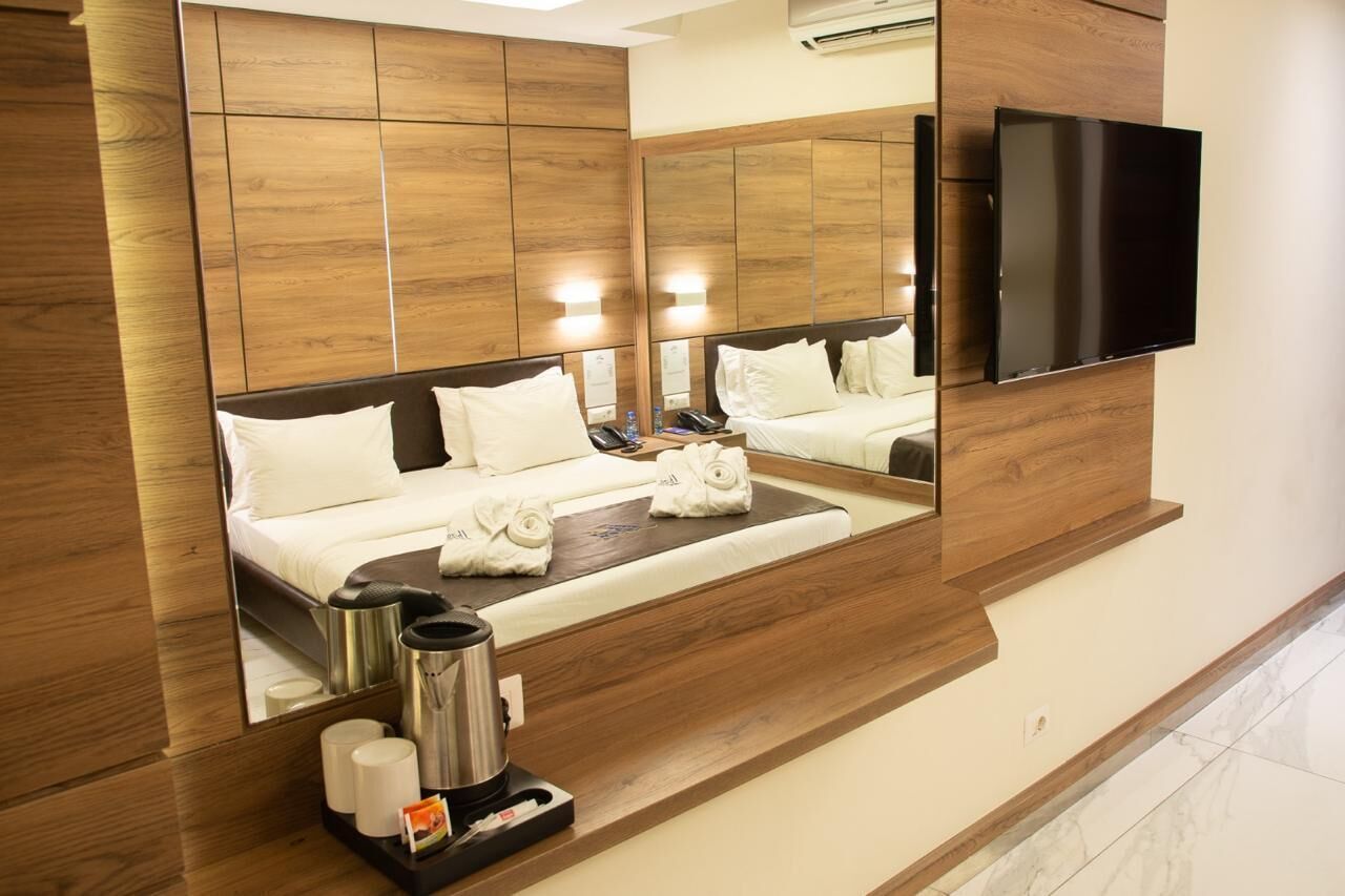 Quarto de hotel com um espelho grande, televisão de tela plana, cama com roupa de cama branca, cafeteira e bandeja com produtos de higiene.