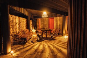 Um quarto com pouca luz, paredes de pedra, uma mesa de madeira e várias cadeiras.