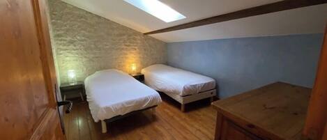 Um quarto pequeno e acolhedor com duas camas de solteiro, piso de madeira, teto inclinado com claraboia e parede de pedra.