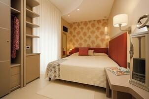 Quarto de hotel com cama, escrivaninha, guarda-roupa e televisão.