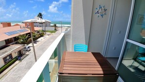 Uma varanda com mesa de madeira e cadeiras azuis, com vista para uma praia e o prédio do Viva Bar.
