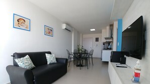 Uma sala de estar moderna com um sofá preto, uma mesa de jantar com cadeiras, uma área de cozinha com televisão e um pequeno objeto decorativo no balcão.