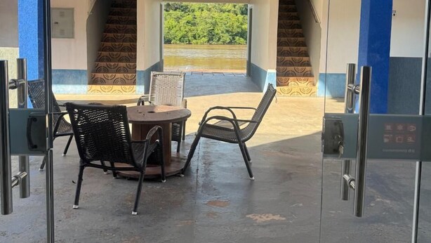 Uma área externa com mesas e cadeiras, uma escadaria que leva a um andar superior e vista para um corpo d'água.