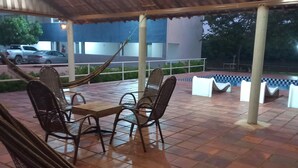Um pátio externo com mesa e cadeiras, uma rede e uma área de piscina ao fundo.