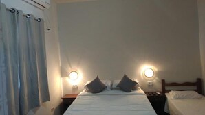 Quarto com uma cama, duas mesas de cabeceira, um criado-mudo, ar-condicionado fixado na parede e uma cortina.