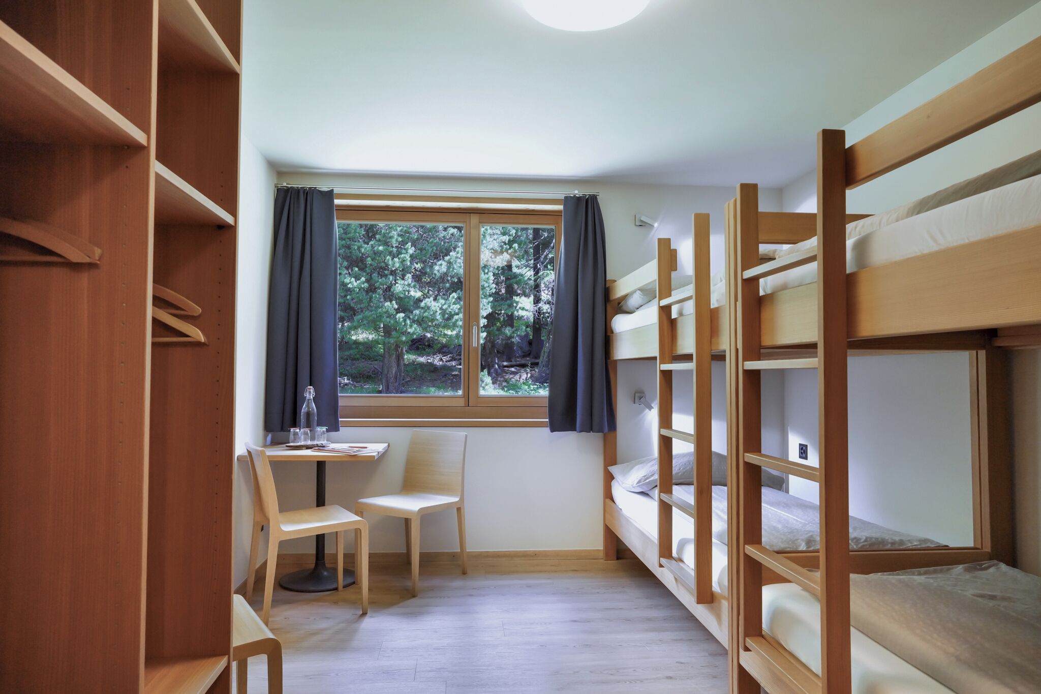 Quarto em dormitório com beliches, uma escrivaninha, duas cadeiras e janela com cortinas.