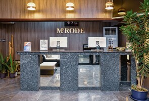 Uma mesa de recepção com uma placa que diz "MRODE", dois monitores Samsung, um relógio digital e uma pequena planta à direita.