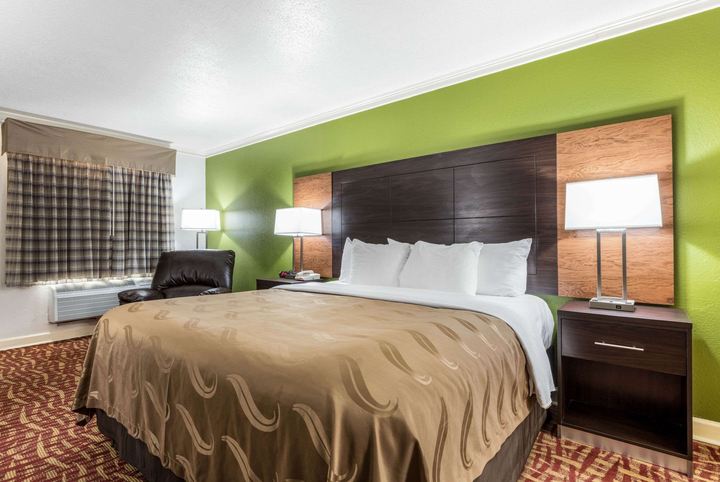 Quarto de hotel com uma cama grande, mesa de cabeceira, cadeira, escrivaninha e janela com cortinas.