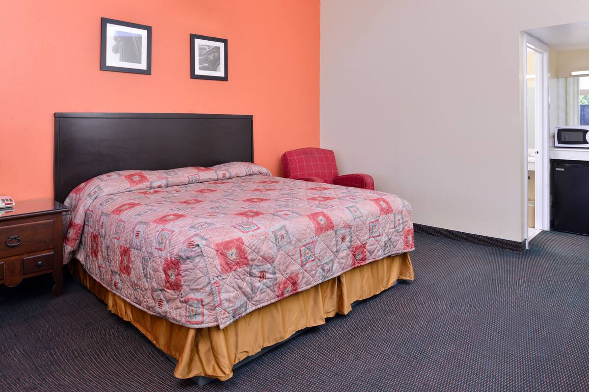 Quarto de hotel com uma cama, uma cadeira vermelha, uma mesa de cabeceira e uma porta que dá para uma cozinha compacta.