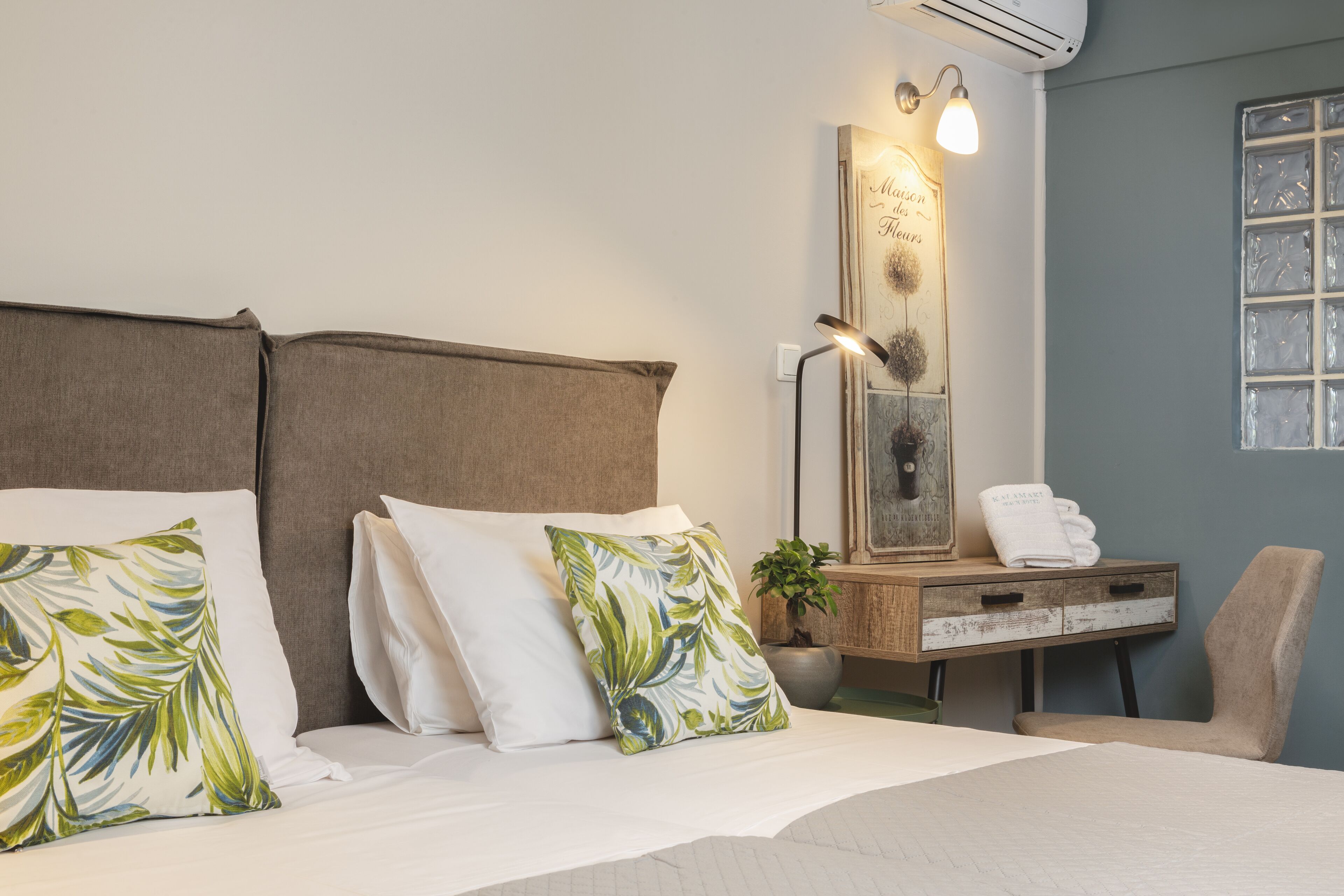 Cama bem arrumada com almofadas brancas e padrão de folhagem tropical, mesa de cabeceira de madeira com luminária, obra de arte emoldurada e uma cadeira.