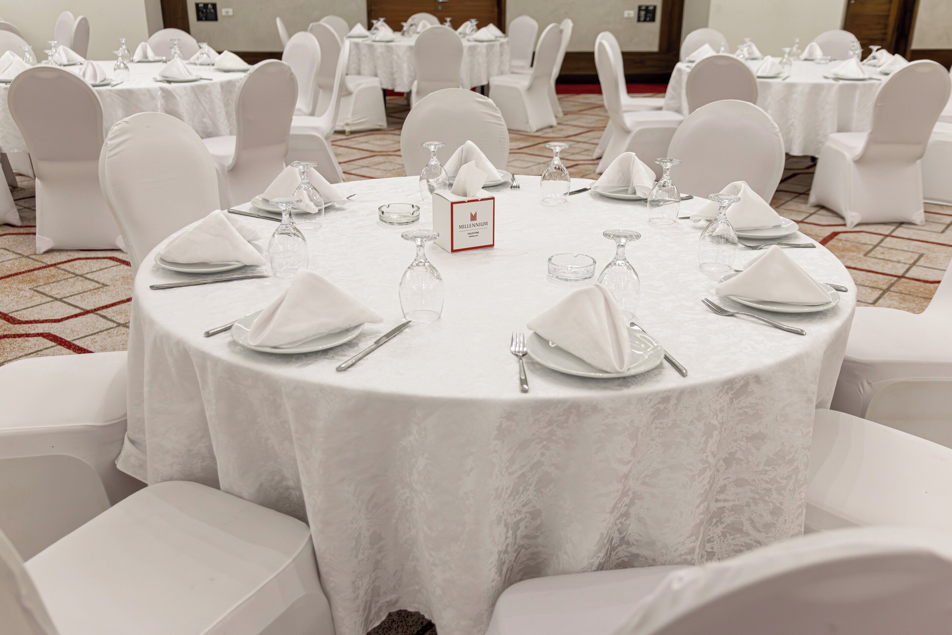 Uma sala de banquetes com mesas redondas preparadas para um evento formal, cada uma com toalhas brancas, copos e guardanapos dobrados.