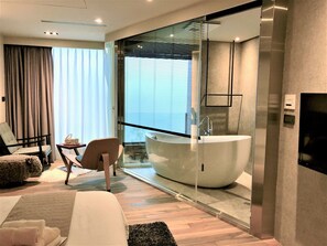 Quarto de hotel moderno com uma cama grande, um espaço de estar com poltrona e mesa, uma banheira espaçosa e um painel de vidro.