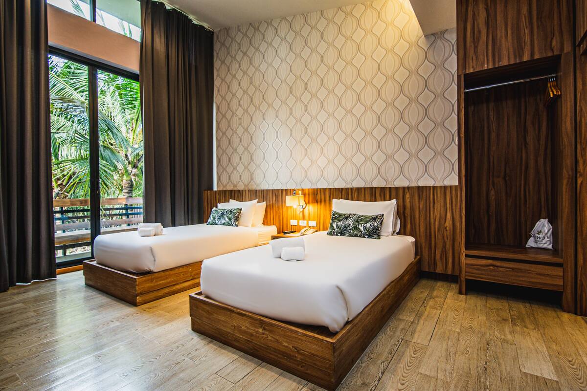 Quarto de hotel com duas camas, piso de madeira, parede estampada e vista para o verde lá fora.