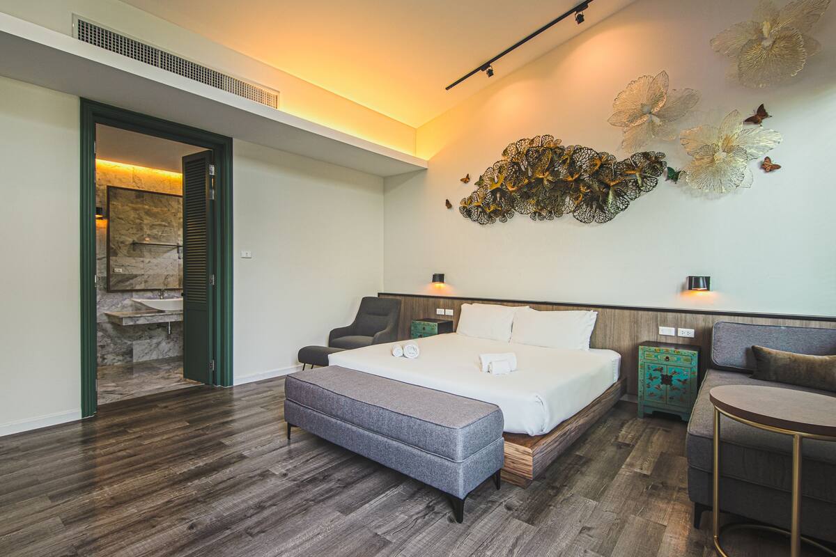 Quarto moderno com uma cama grande, um banquinho, uma mesinha e um candeeiro fixado na parede.