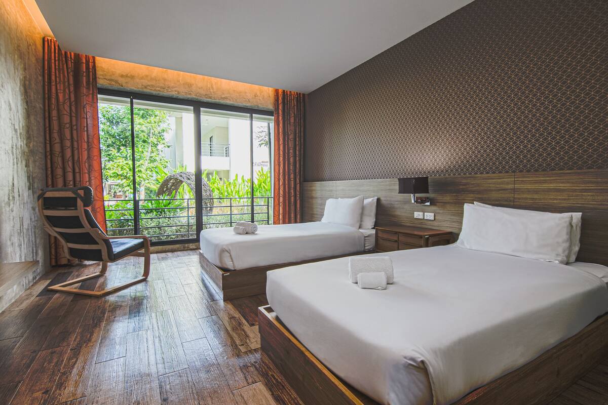 Quarto de hotel com duas camas, uma cadeira de balanço, uma janela grande com cortinas e uma parede estampada.