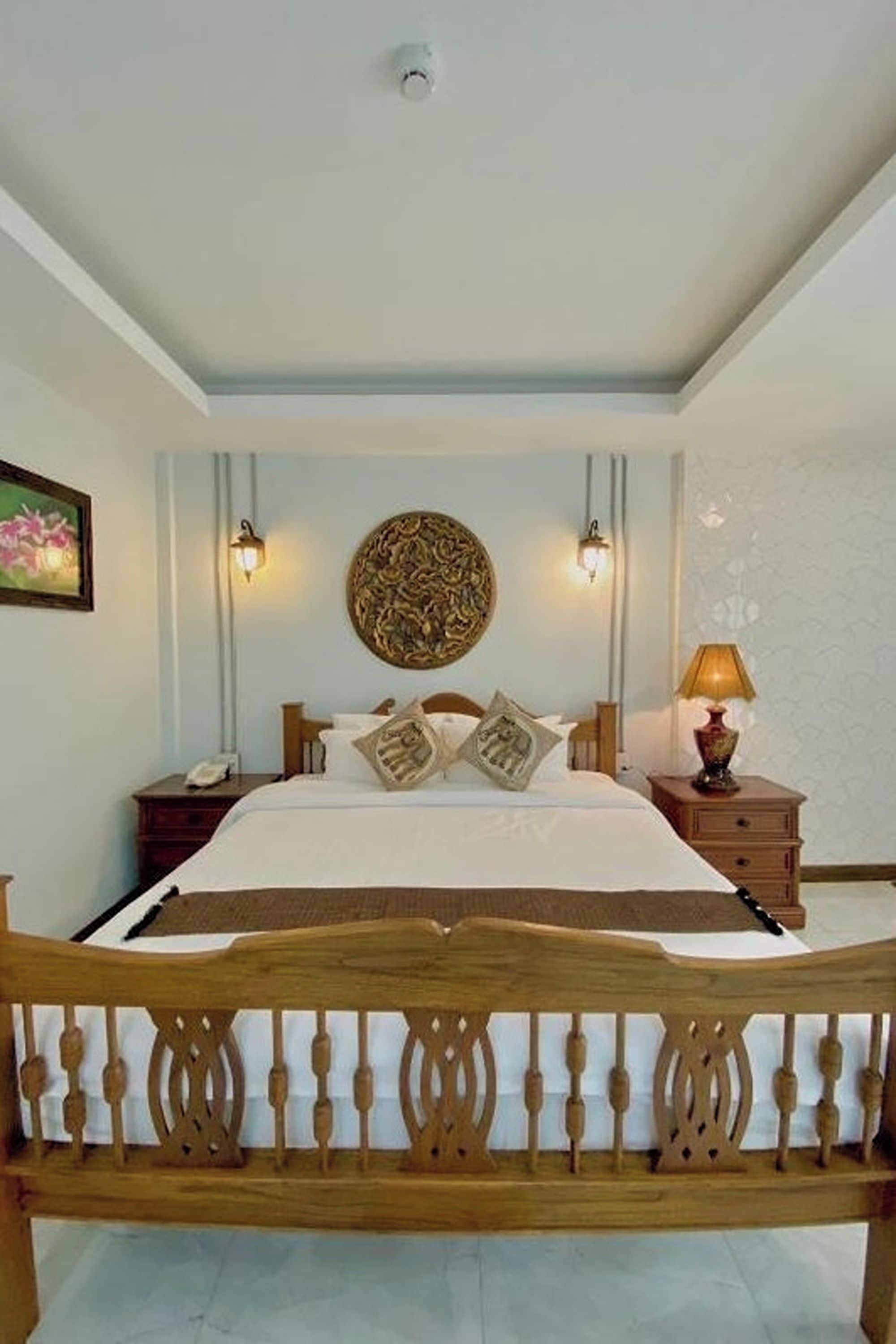 Quarto com cama de madeira, duas mesas de cabeceira, luminária na parede e um objeto decorativo na parede.