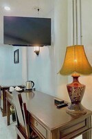 Quarto de hotel com televisão de tela plana na parede, mesa com luminária, chaleira e controle remoto.