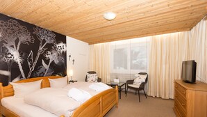 Quarto de hotel com cama de madeira, uma mesa com cadeira, televisão sobre um armário de madeira e uma grande janela com cortinas.