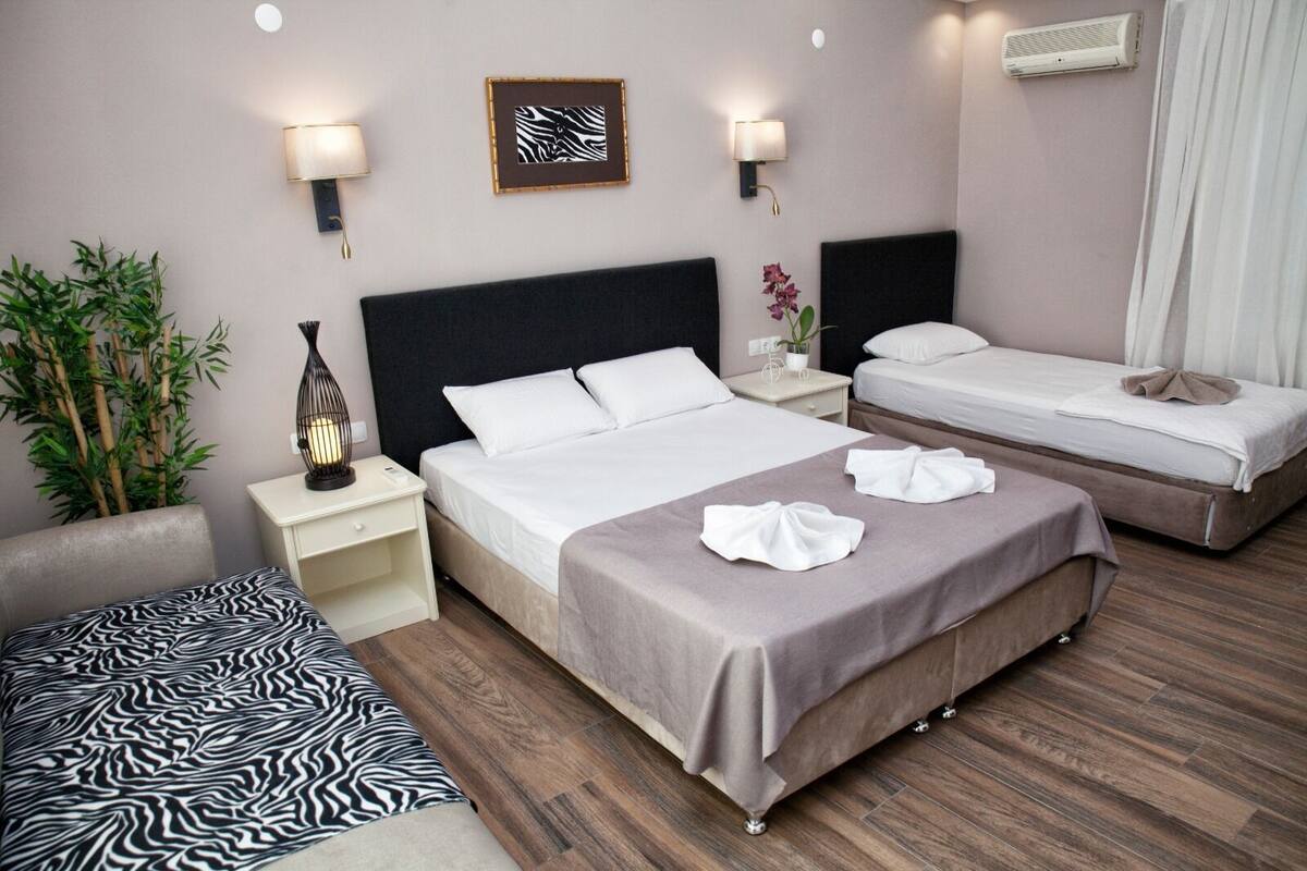 Quarto de hotel com uma cama grande, uma cama menor, mesa de cabeceira com abajur, planta em vaso e ar condicionado fixado na parede.