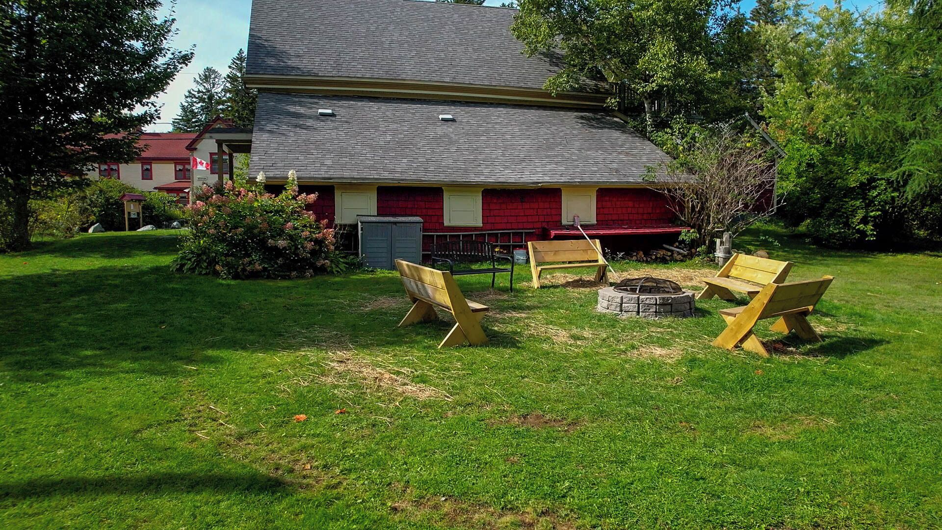 Uma casa de tijolos vermelhos com telhado cinzento, um poço de fogo e duas bancos de madeira num jardim gramado.