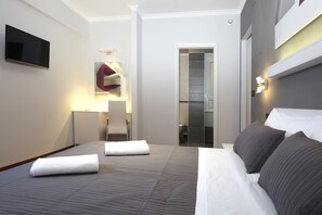 Quarto de hotel com cama, televisão, escrivaninha e banheiro visível através de uma porta aberta.
