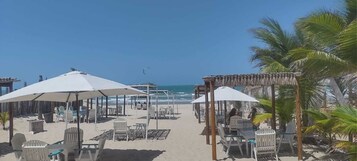 Uma praia com fileiras de cadeiras e mesas brancas sob guarda-sóis brancos, uma estrutura de palha e palmeiras.