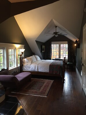 Quarto com teto inclinado, uma cama, uma cadeira, uma mesa e uma janela com cortinas.