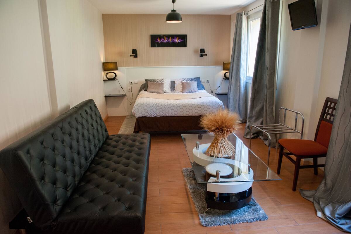 Quarto de hotel moderno com uma cama, um sofá preto, uma mesinha com um vaso decorativo e uma cadeira.