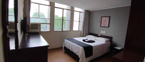 Quarto de hotel com cama, escrivaninha e janela com vista para o verde.