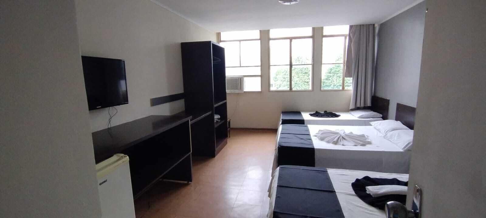 Quarto de hotel com cama, escrivaninha, televisão e janela com cortinas.