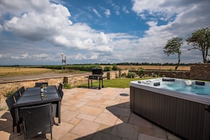 Um pátio com hidromassagem, mesa e cadeiras externos, cercado por um muro de tijolos e vista para um campo.