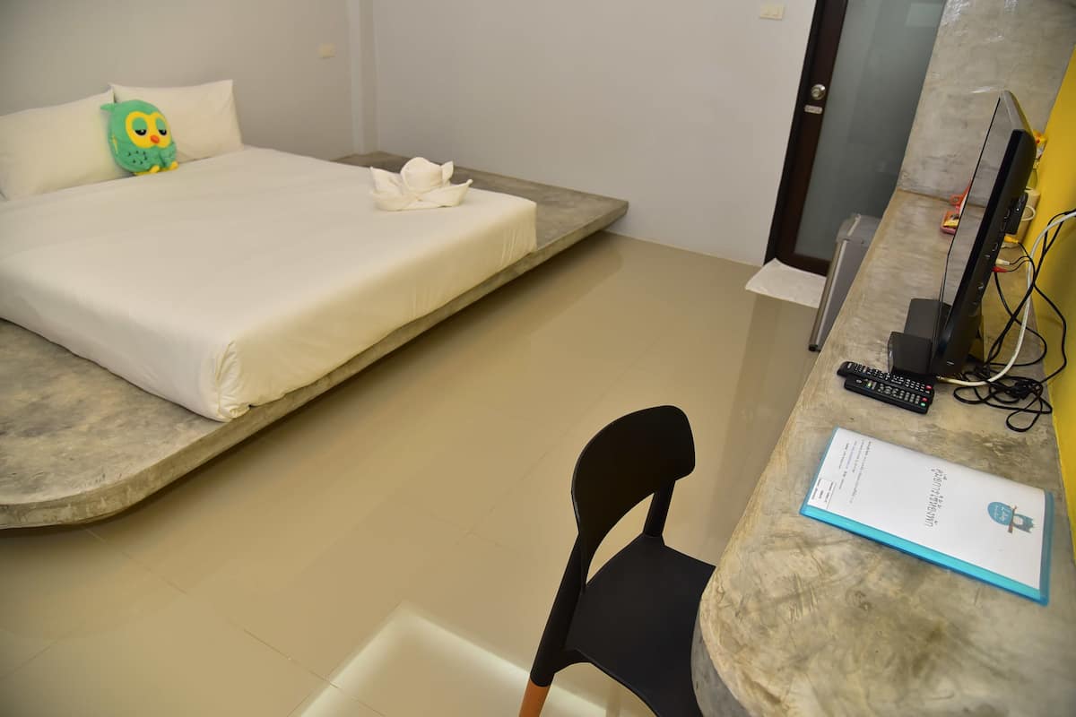 Uma cama com travesseiro e um brinquedo de pelúcia, uma mesa com computador e controle remoto, e uma cadeira.