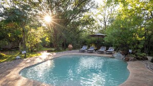 Piscina com espreguiçadeiras e um guarda-sol, rodeada por árvores e um deck de pedra.