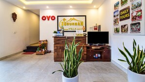 Uma área de recepção com uma placa indicando "Suburban Villa" e "OYO". Há plantas em vasos, um balcão de recepção e uma televisão.