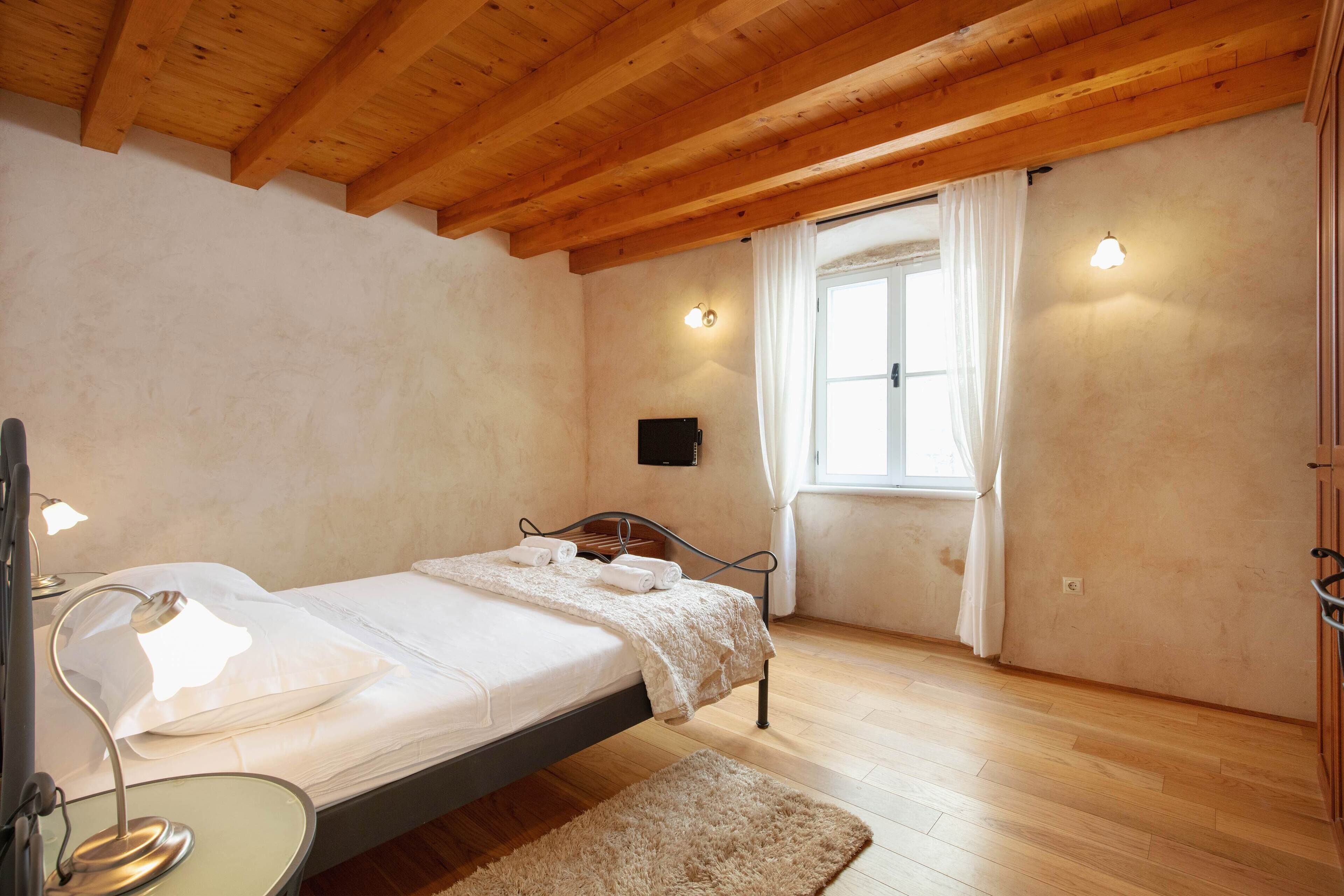 Quarto com teto de madeira, cama com roupa branca, abajur e janela com cortinas.