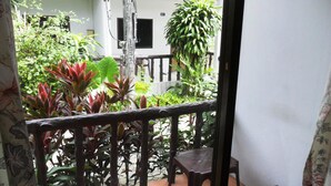 Uma varanda com guarda-corpo de madeira, rodeada por diversas plantas verdes e vista para um edifício com janela e porta.