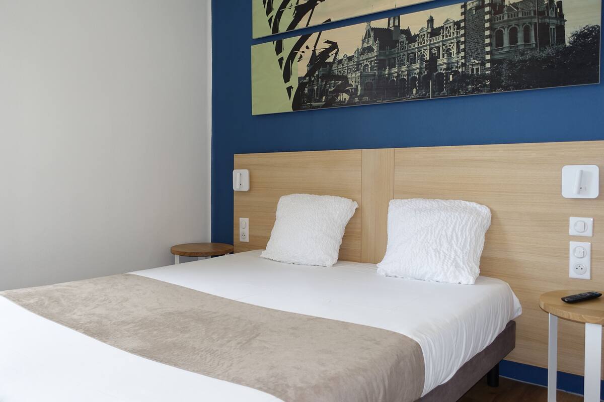 Quarto de hotel com uma cama, uma mesa de cabeceira, um interruptor de luz na parede e um quadro sobre a cama.