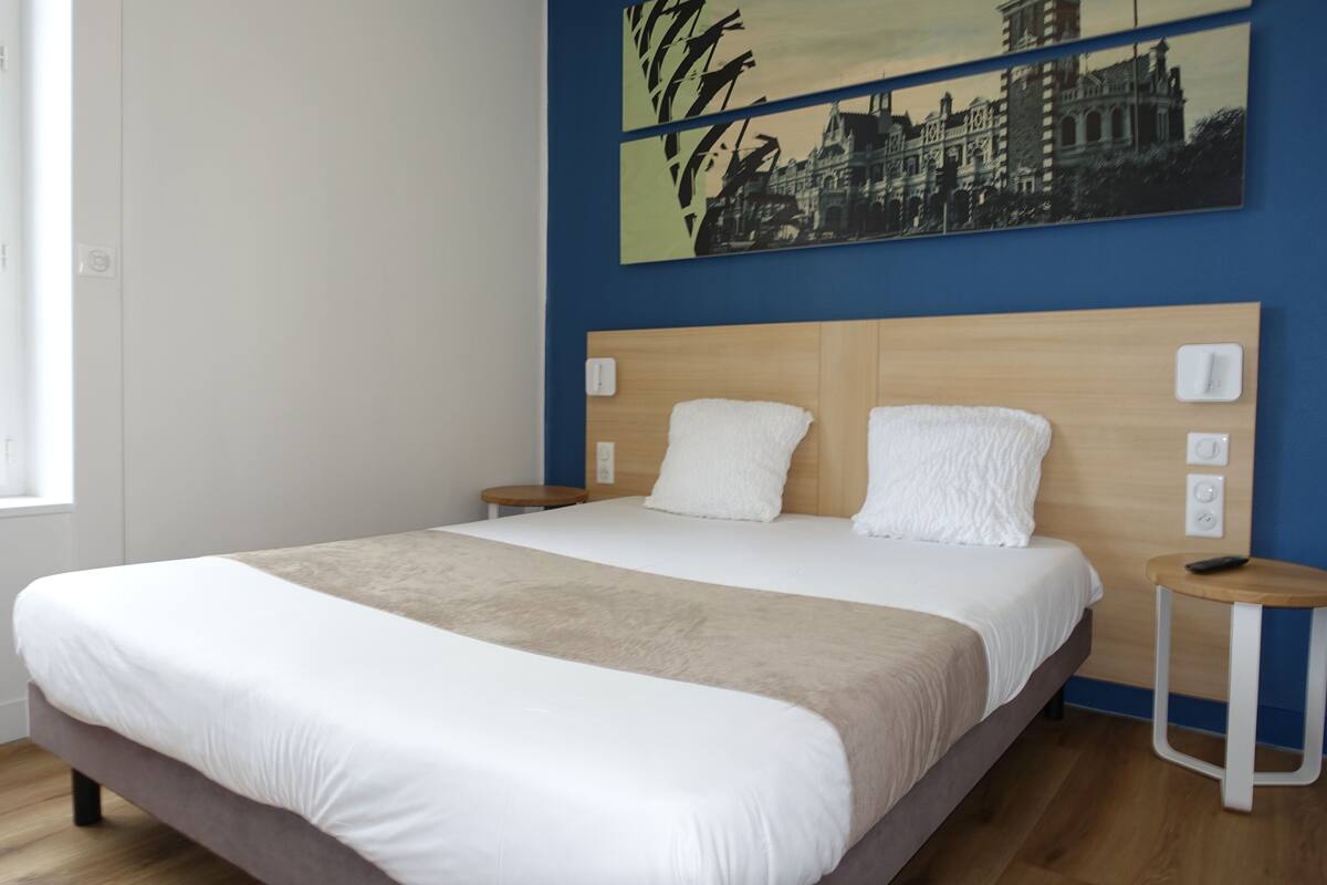 Uma cama bem arrumada com cabeceira de madeira, uma mesa de cabeceira com um controle remoto e um interruptor de luz na parede.