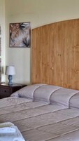 Quarto com cabeceira de madeira, cama com colcha em tons claros, mesa de cabeceira com abajur e quadro na parede com folhas tropicais.