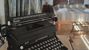 Uma máquina de escrever antiga sobre uma mesa em quarto de hotel, com uma cama e cortinas ao fundo.