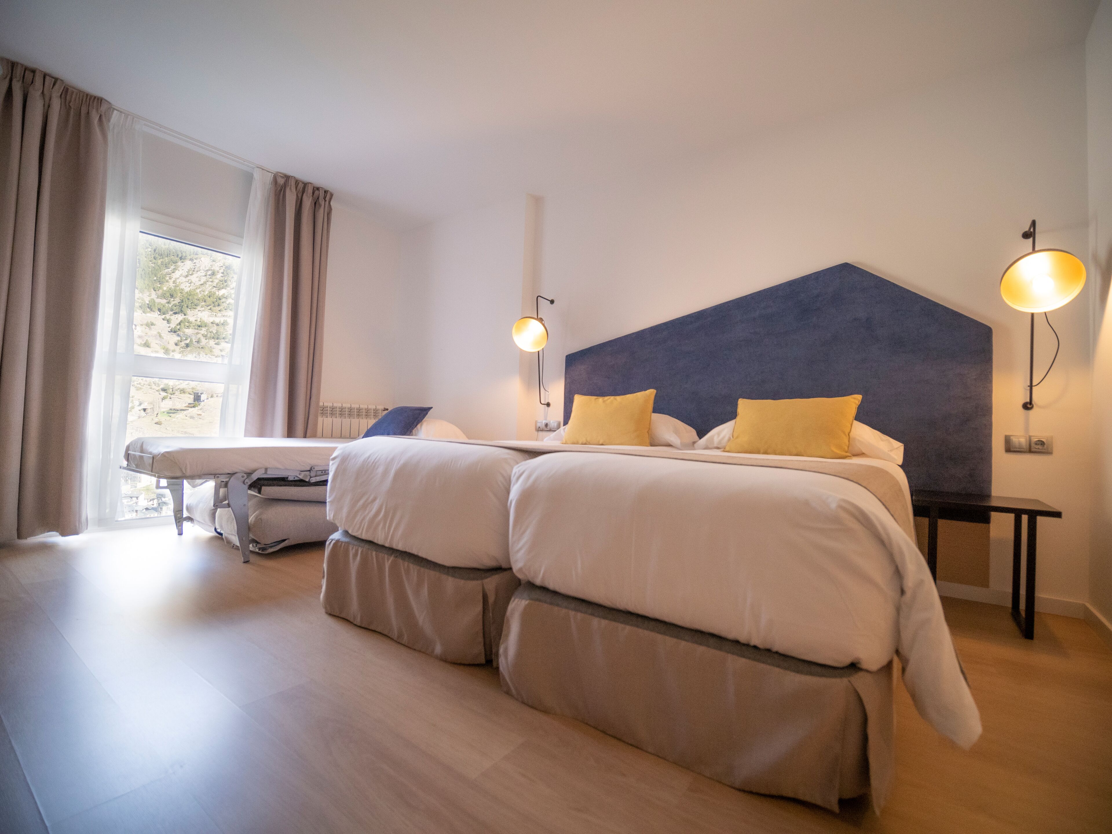 Quarto de hotel moderno com uma cama grande, mesas de cabeceira e vista para uma paisagem montanhosa pela janela.