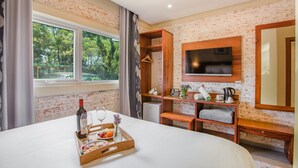 Quarto de hotel com uma cama grande, televisão fixada na parede, prateleira de madeira com telefone e um vaso, e janela com vista para o verde.