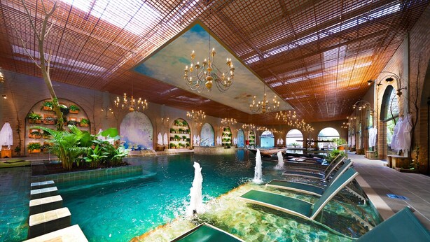 Uma área interna de piscina luxuosa, com várias áreas de estar, uma fonte central e um teto com padrão em grade.