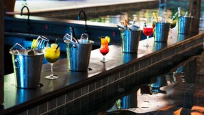 Uma área de bar com bebidas em baldes e copos de metal, um balcão revestido com azulejos e uma piscina ao fundo.