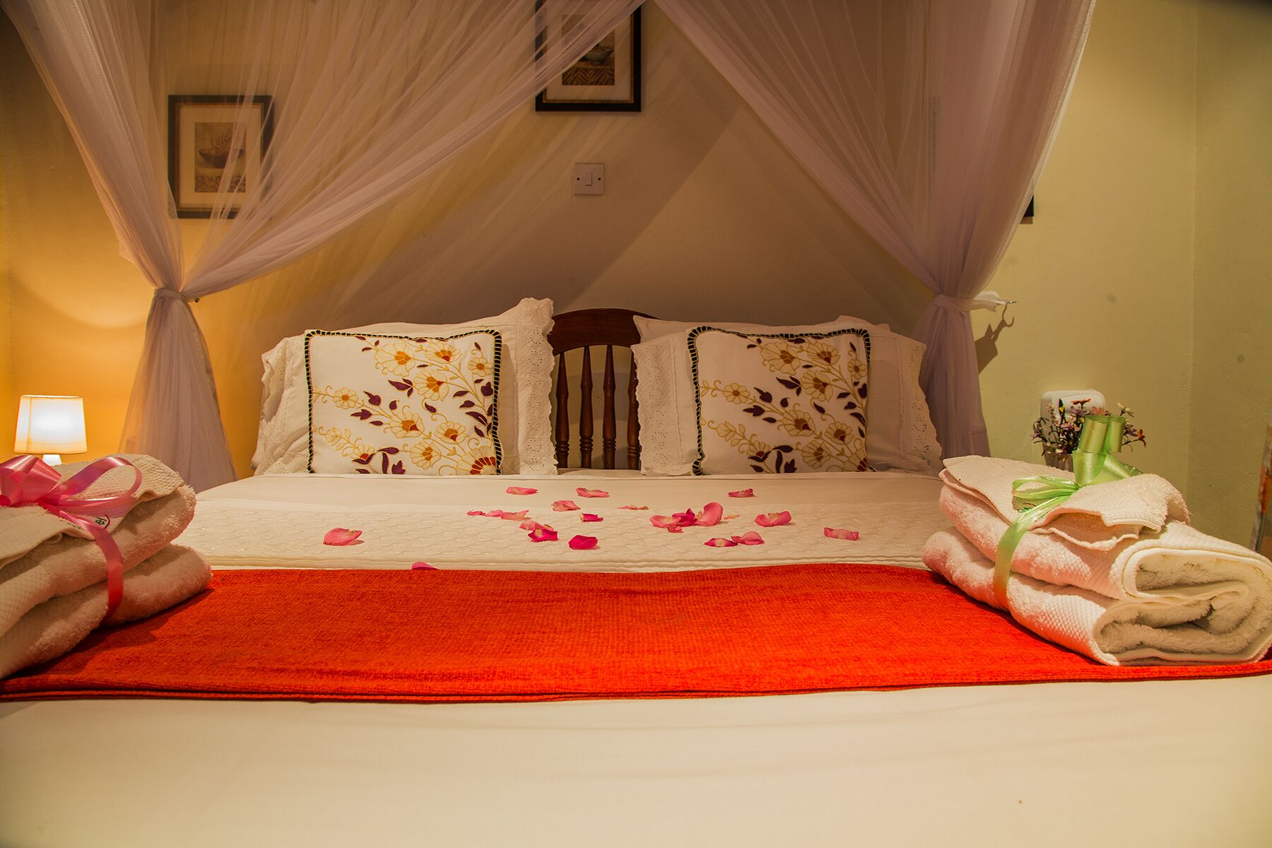 Cama com manta vermelha, almofadas florais e pétalas de rosa. Há toalhas e um vaso com flores ao lado.