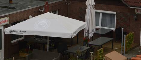 Uma área externa com mesas e cadeiras, coberta por um grande guarda-sol branco, e uma placa para Franziskaner Weissbier.