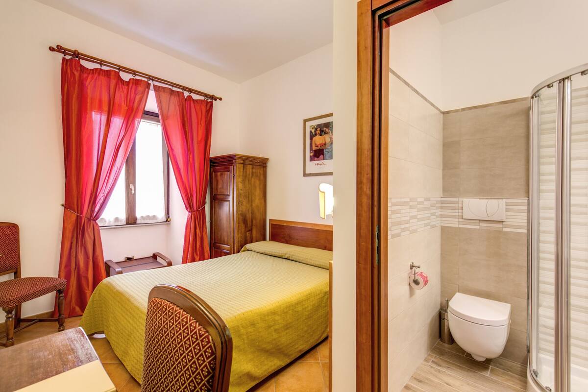 Quarto de hotel com cama, cadeira, armário de madeira, janela com cortinas vermelhas e banheiro com vaso sanitário e chuveiro.